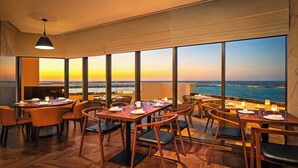 Restaurante com grandes janelas oferecendo vista para o mar ao pôr do sol. Mesas montadas com cadeiras e velas.