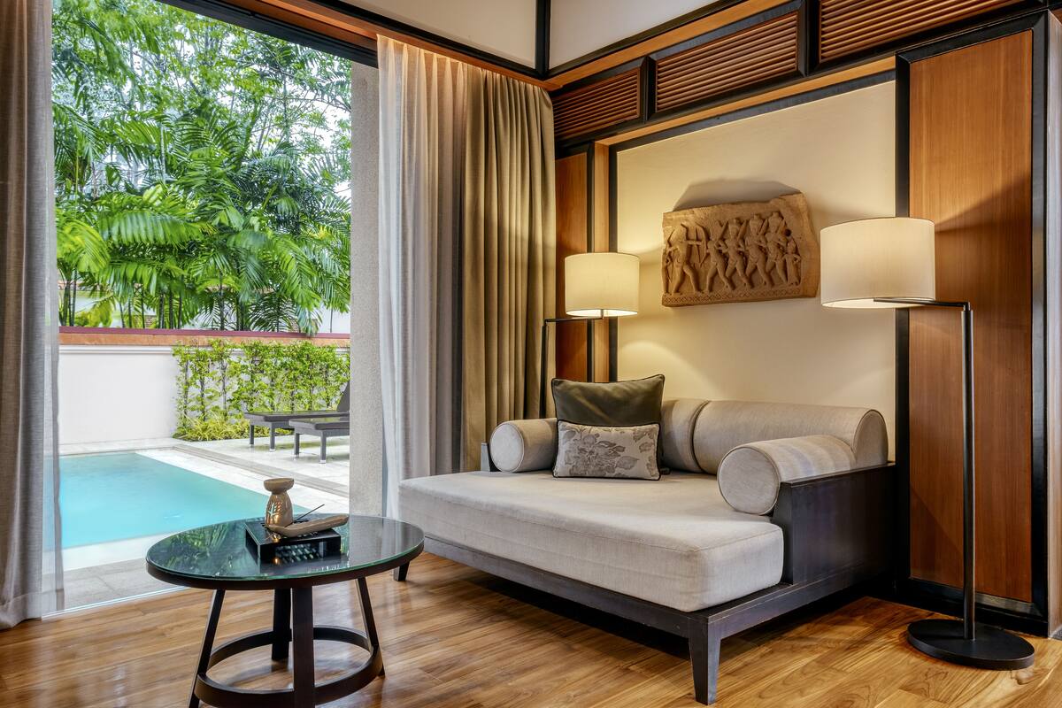 Quarto de hotel moderno com sofá, duas luminárias de chão, uma mesa pequena e vista para uma piscina e áreas verdes lá fora.