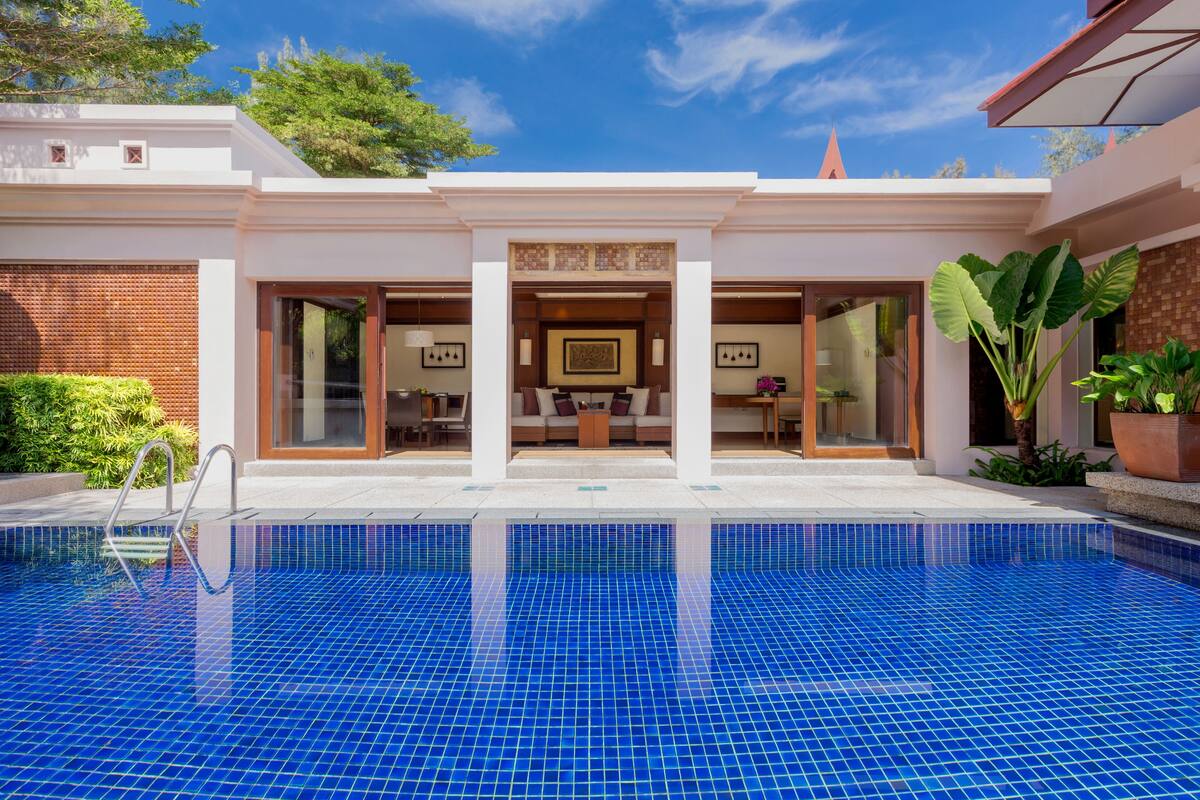 Uma área de piscina moderna com uma piscina azul clara e ladrilhada, um espaço de estar coberto com um sofá e mesa, e uma parede de tijolos em um dos lados.