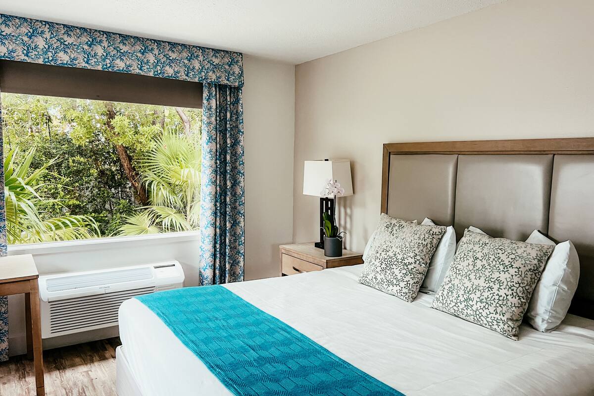 Quarto de hotel com uma cama, mesa de cabeceira, luminária e janela com cortinas.