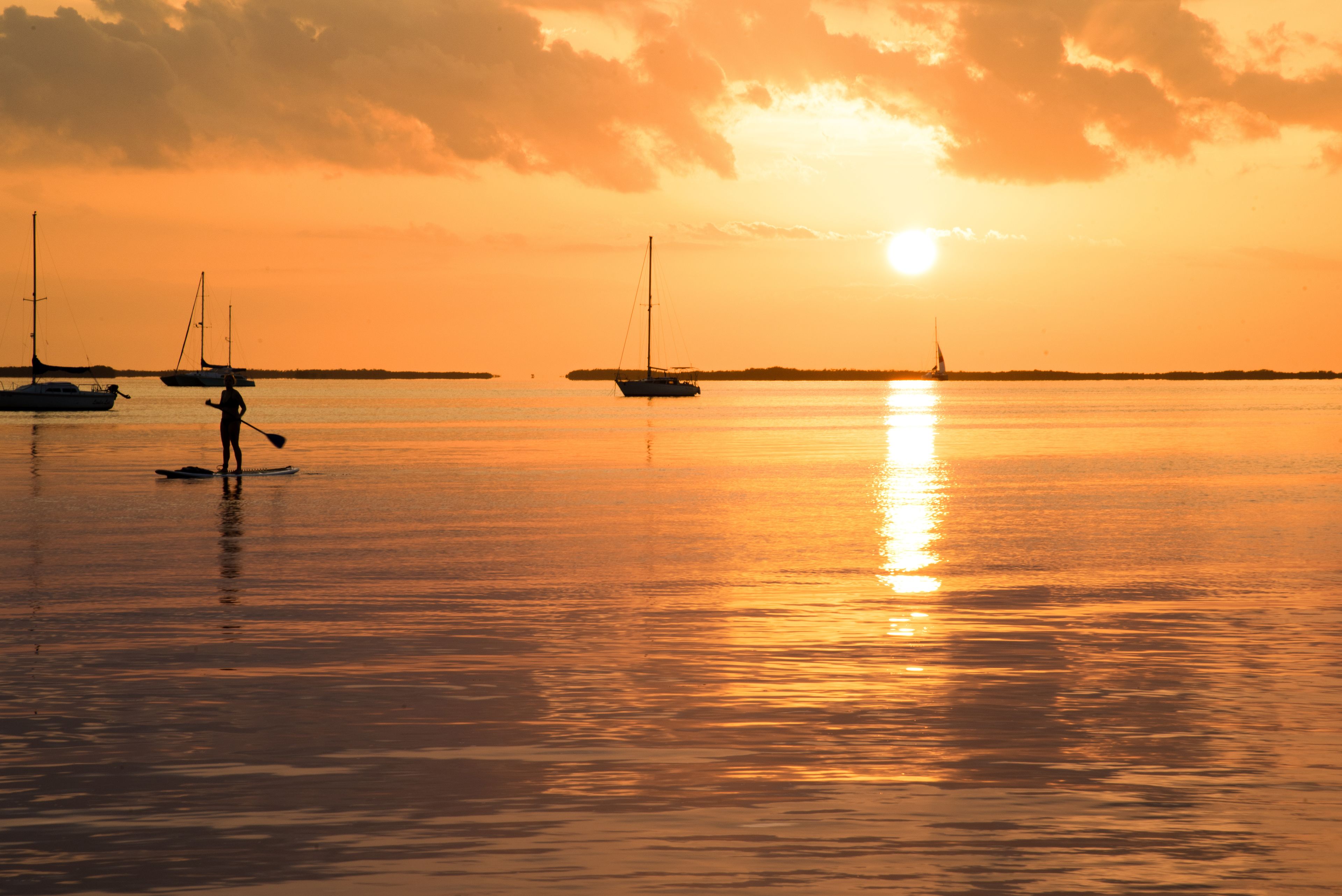 Pôr do sol sobre águas calmas com barcos e uma pessoa praticando paddleboard.