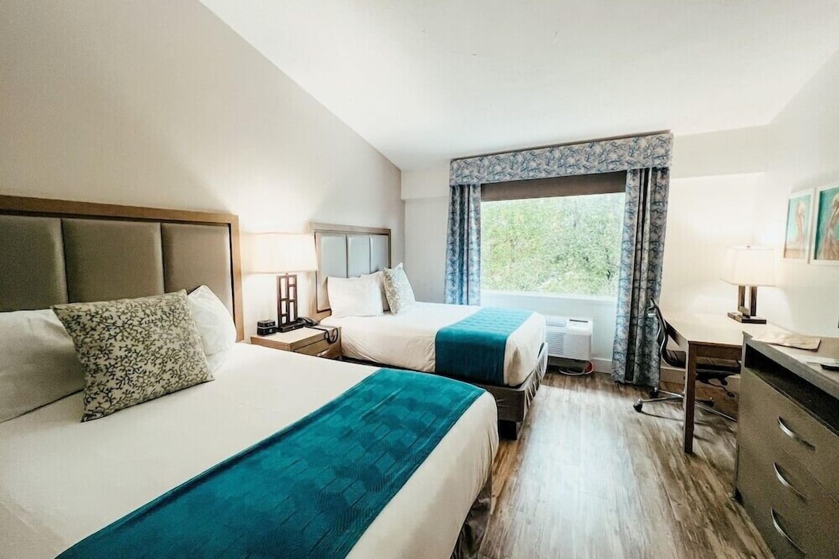 Quarto de hotel com duas camas, uma escrivaninha e uma janela com cortinas.