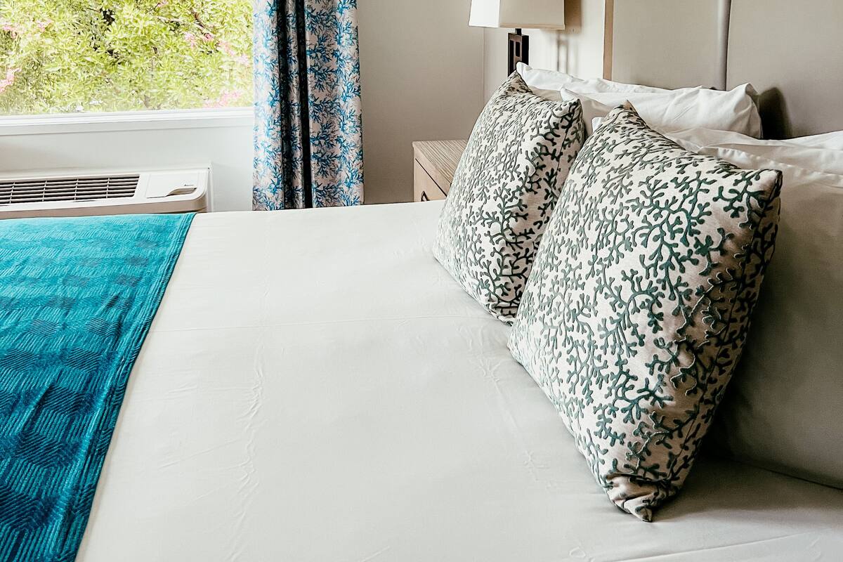 Quarto de hotel com uma cama grande, janela com cortinas, mesa de cabeceira com abajur e vista para o verde lá fora.