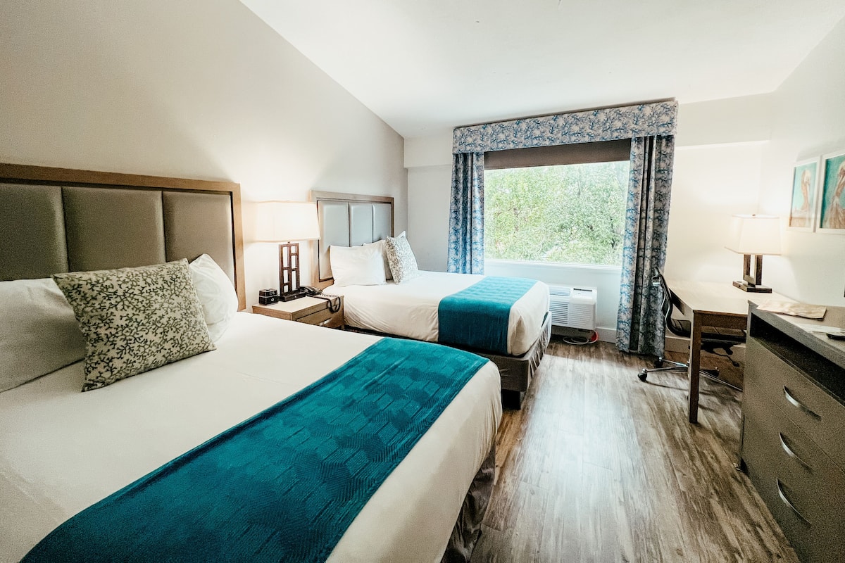 Quarto de hotel com duas camas, uma escrivaninha, uma cadeira e uma janela com cortinas.
