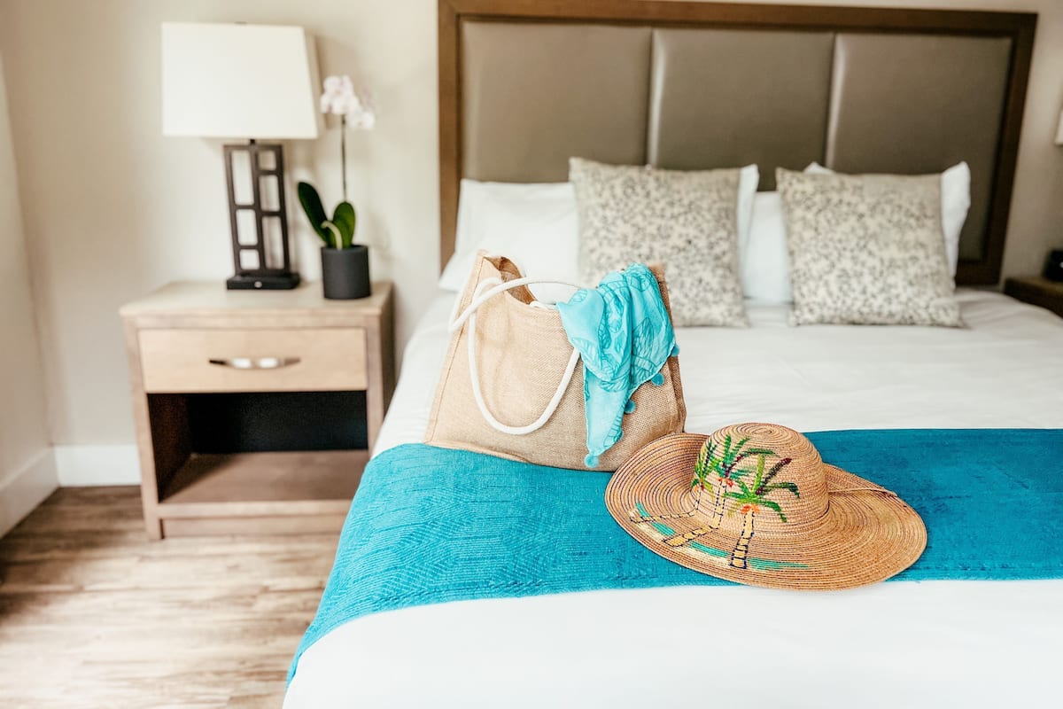 Uma cama bem arrumada com um edredom azul, um chapéu de vime e uma bolsa bege ao lado.