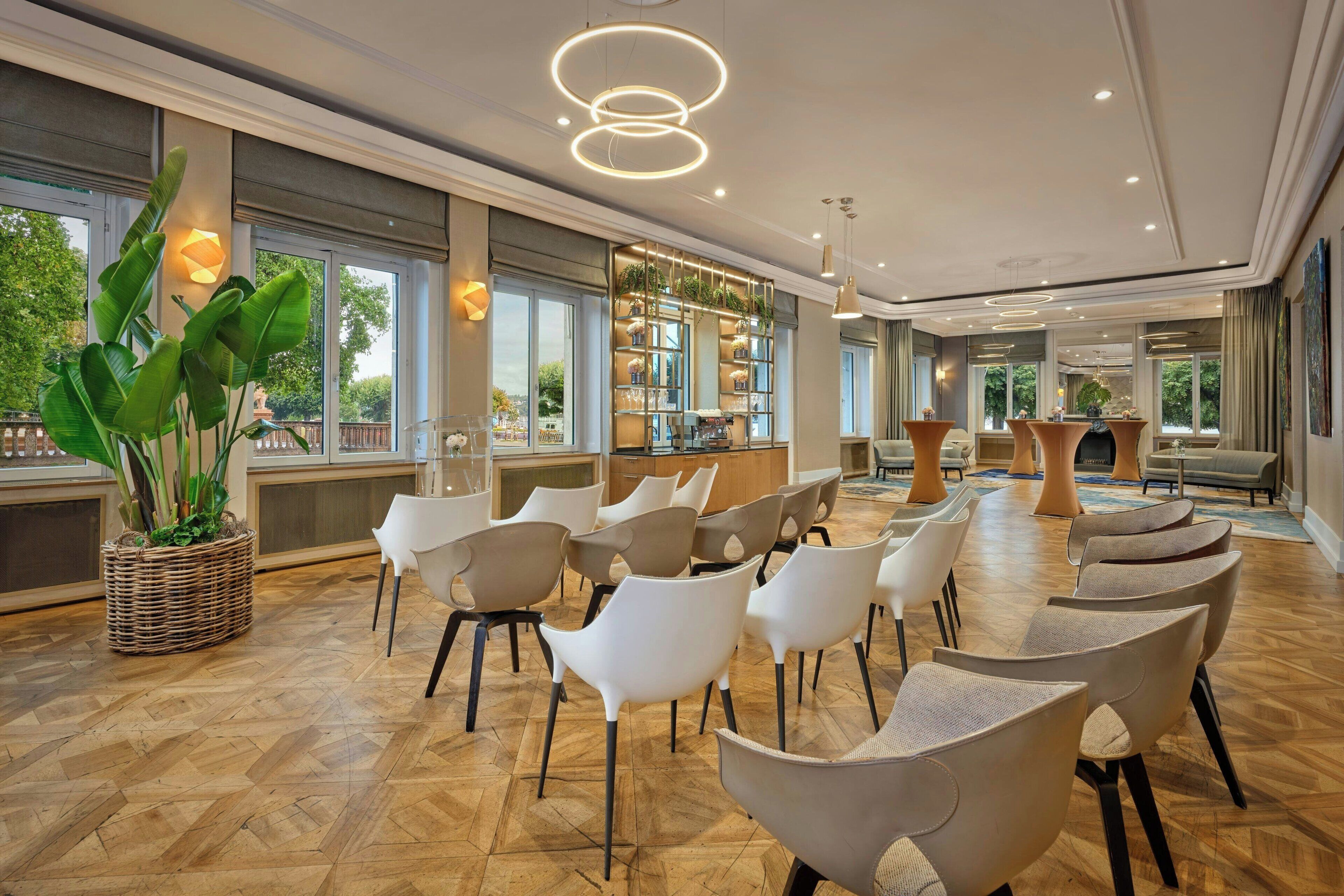 Uma área de estar ampla e bem iluminada, com cadeiras modernas, uma grande planta em vaso e um bar com prateleiras cheias de copos e garrafas.