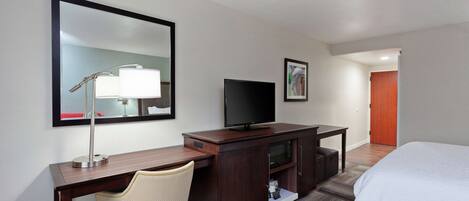 Quarto de hotel com cama, escrivaninha com cadeira, televisão, espelho e porta que dá para outro aposento.