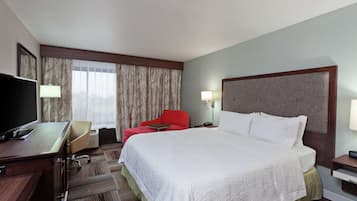 Quarto de hotel com uma cama grande, uma escrivaninha com televisão de tela plana, uma cadeira vermelha e uma janela com cortinas.