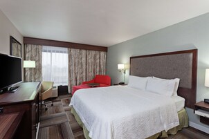 Quarto de hotel com uma cama grande, uma escrivaninha com televisão de tela plana, uma cadeira vermelha e uma janela com cortinas.