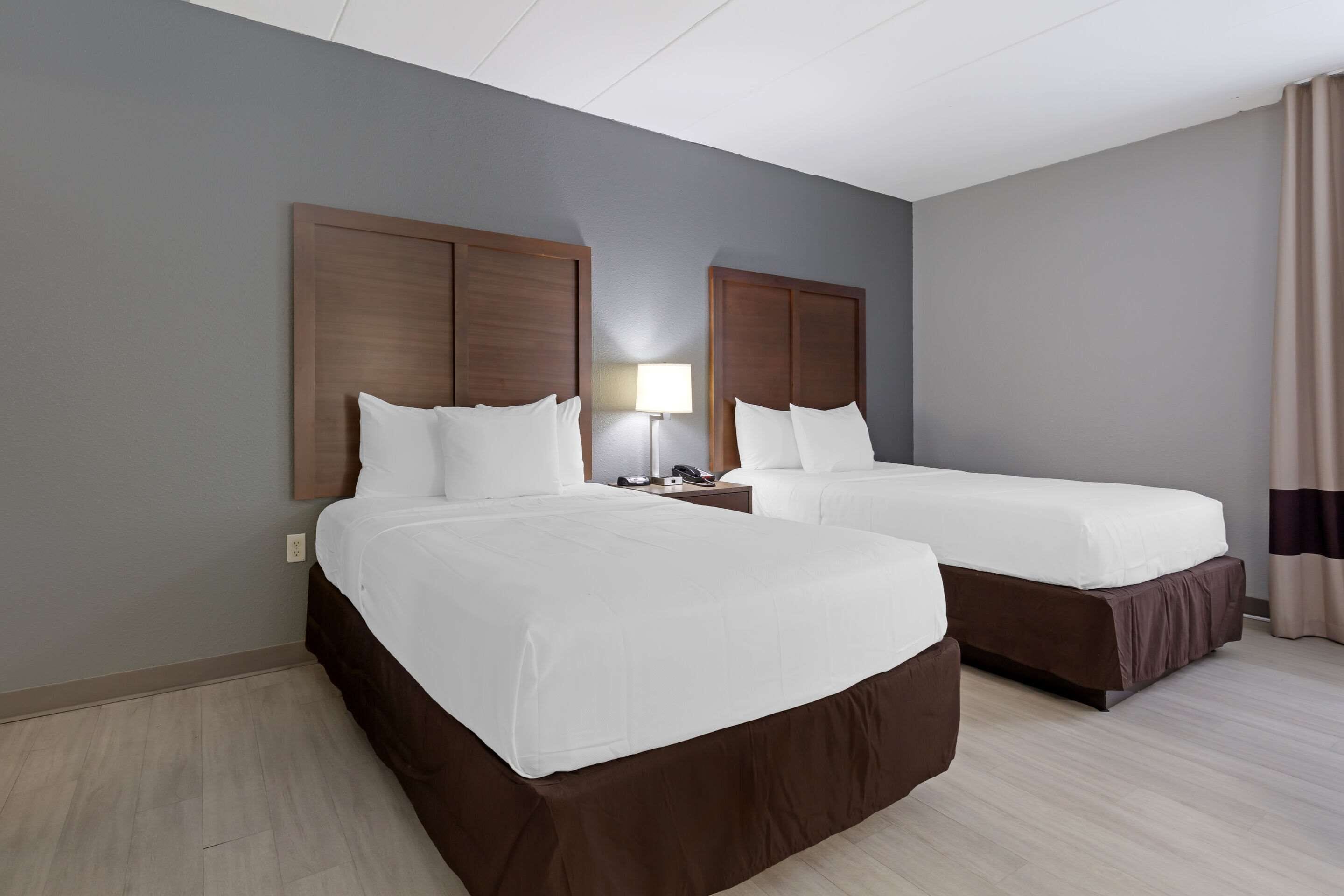 Quarto de hotel com duas camas, mesa de cabeceira, abajur e janela com cortinas.