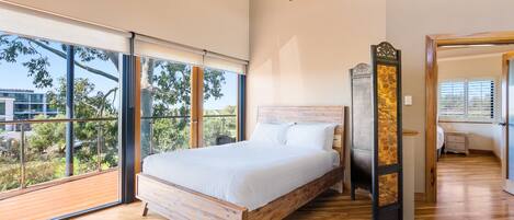 Quarto com cama grande, piso de madeira, vista para área verde e luminária no teto.