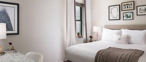 Quarto de hotel com cama, escrivaninha, cadeira, janela com cortinas e quadros na parede.