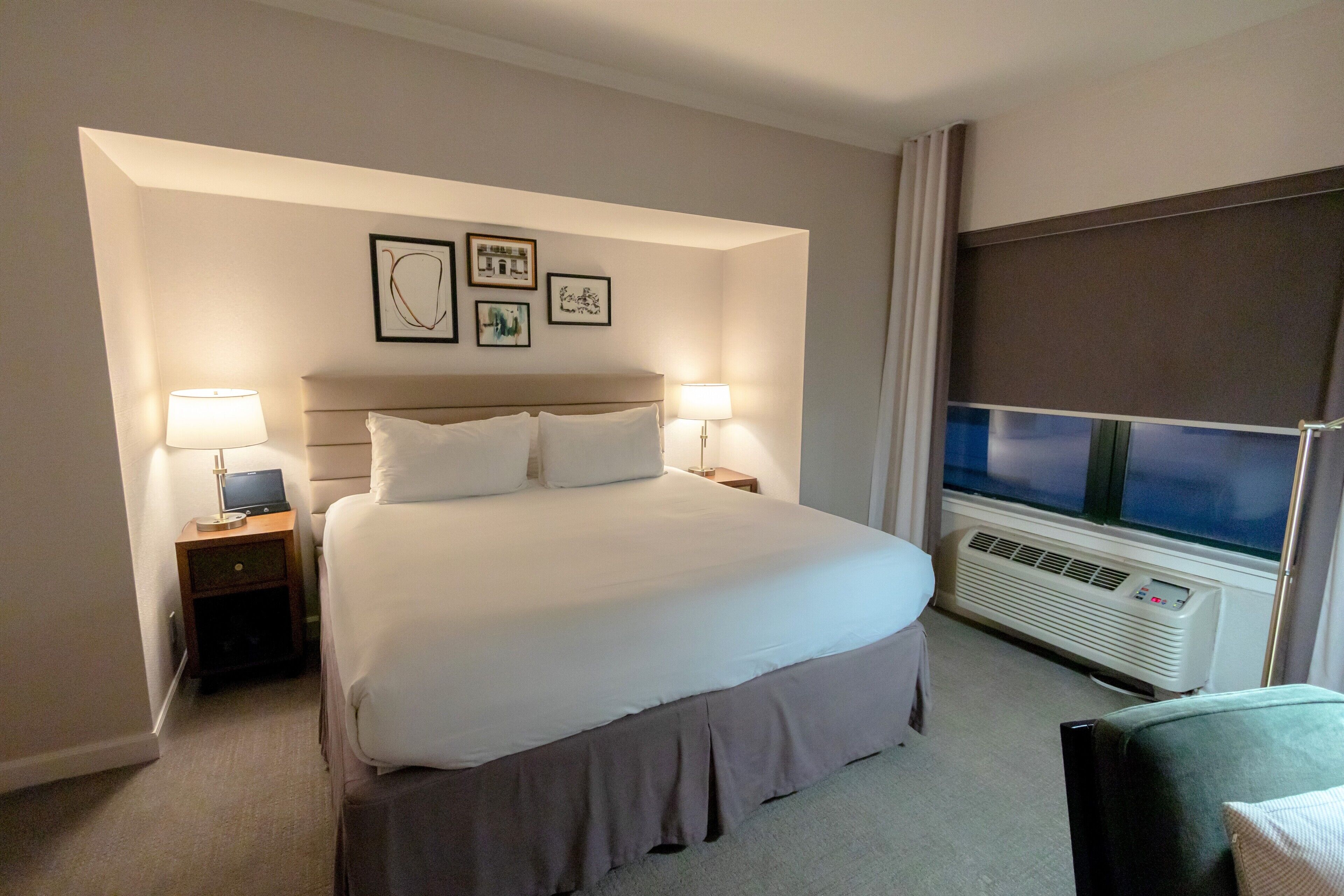 Quarto de hotel com uma cama grande, duas mesas de cabeceira com luminárias, uma janela com vista e uma parede com quadros.