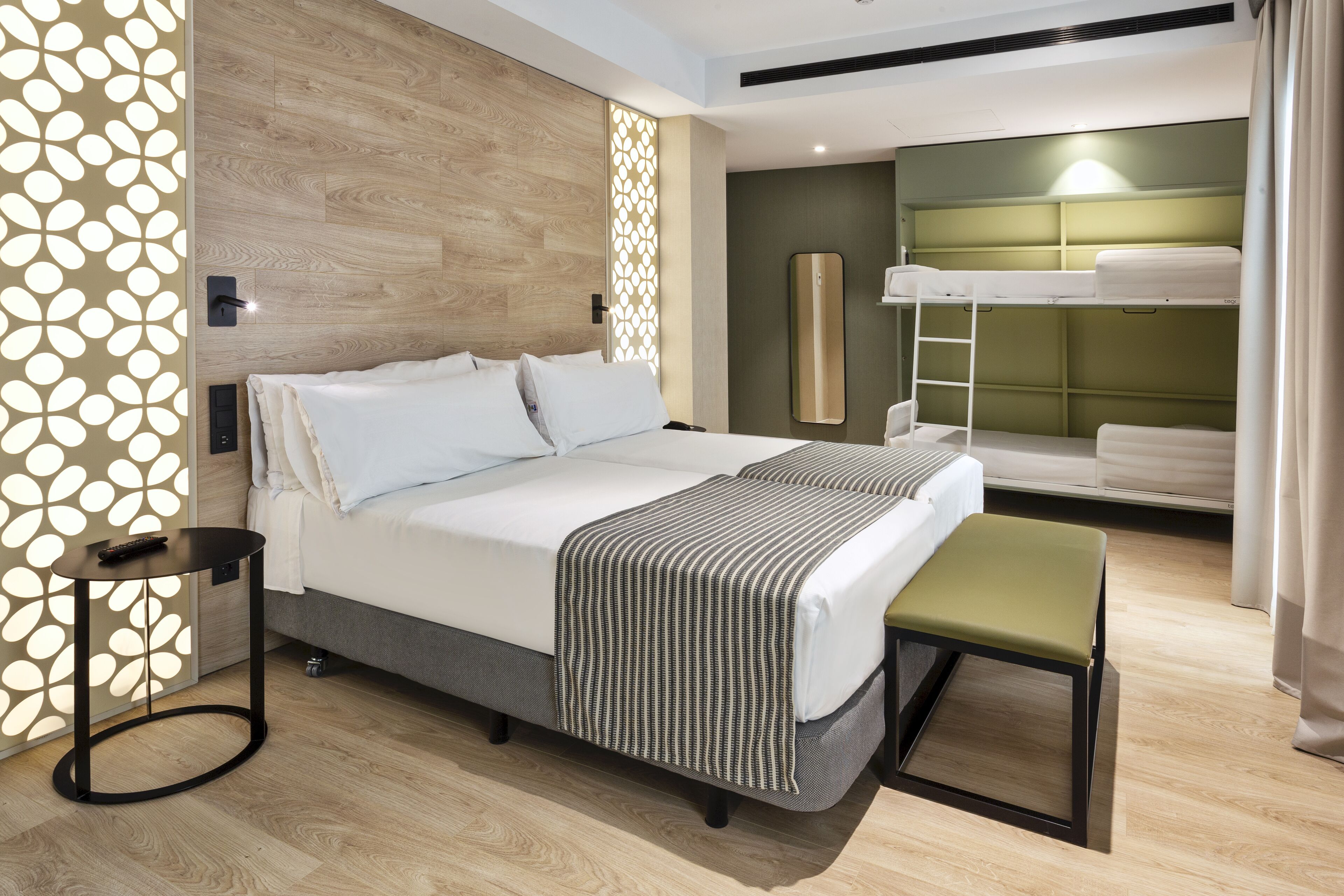 Quarto de hotel moderno com cama, mesa de cabeceira, um banco e uma cama beliche ao fundo.