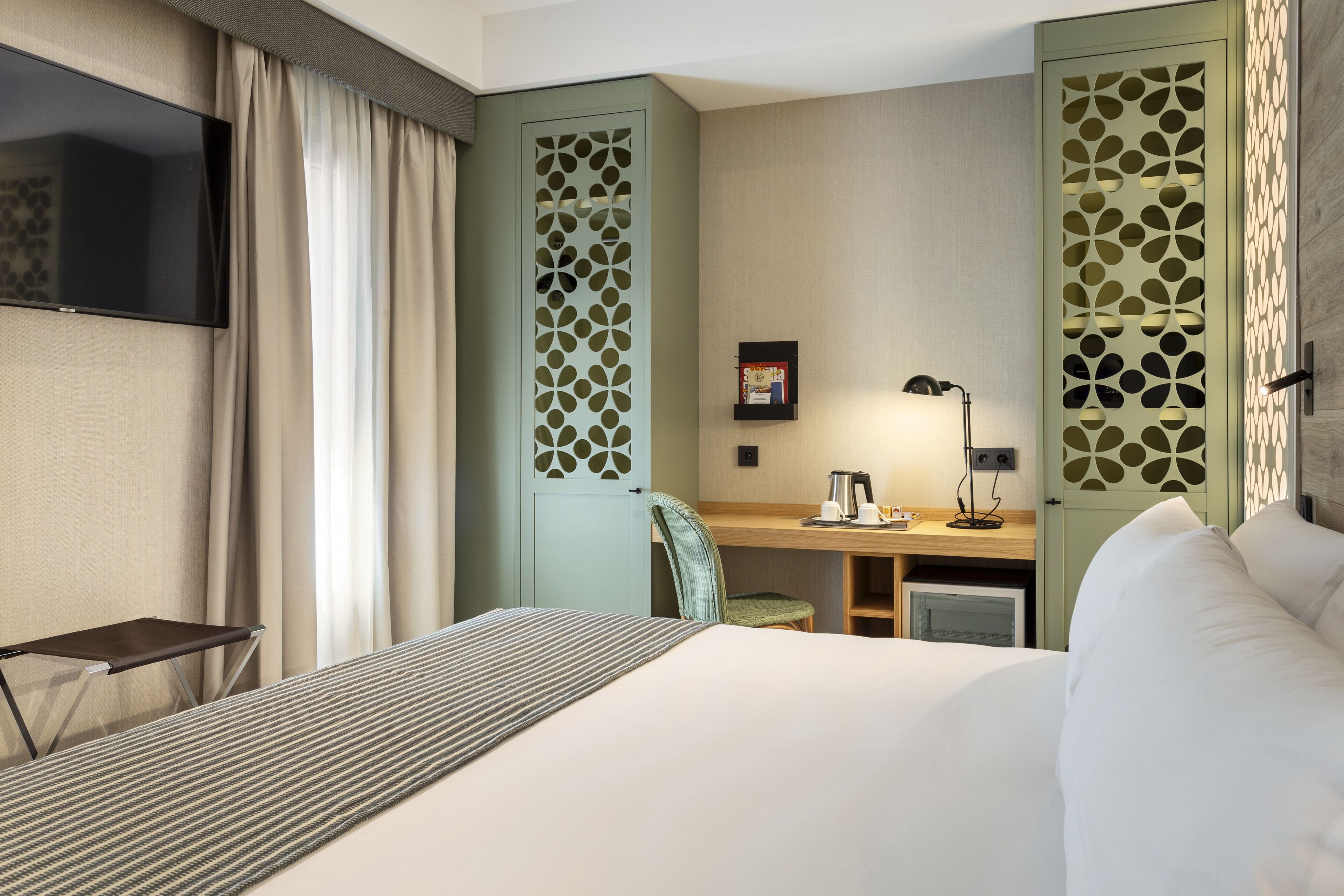 Quarto de hotel com uma cama, uma secretária com cadeira, uma televisão na parede e um minibar.
