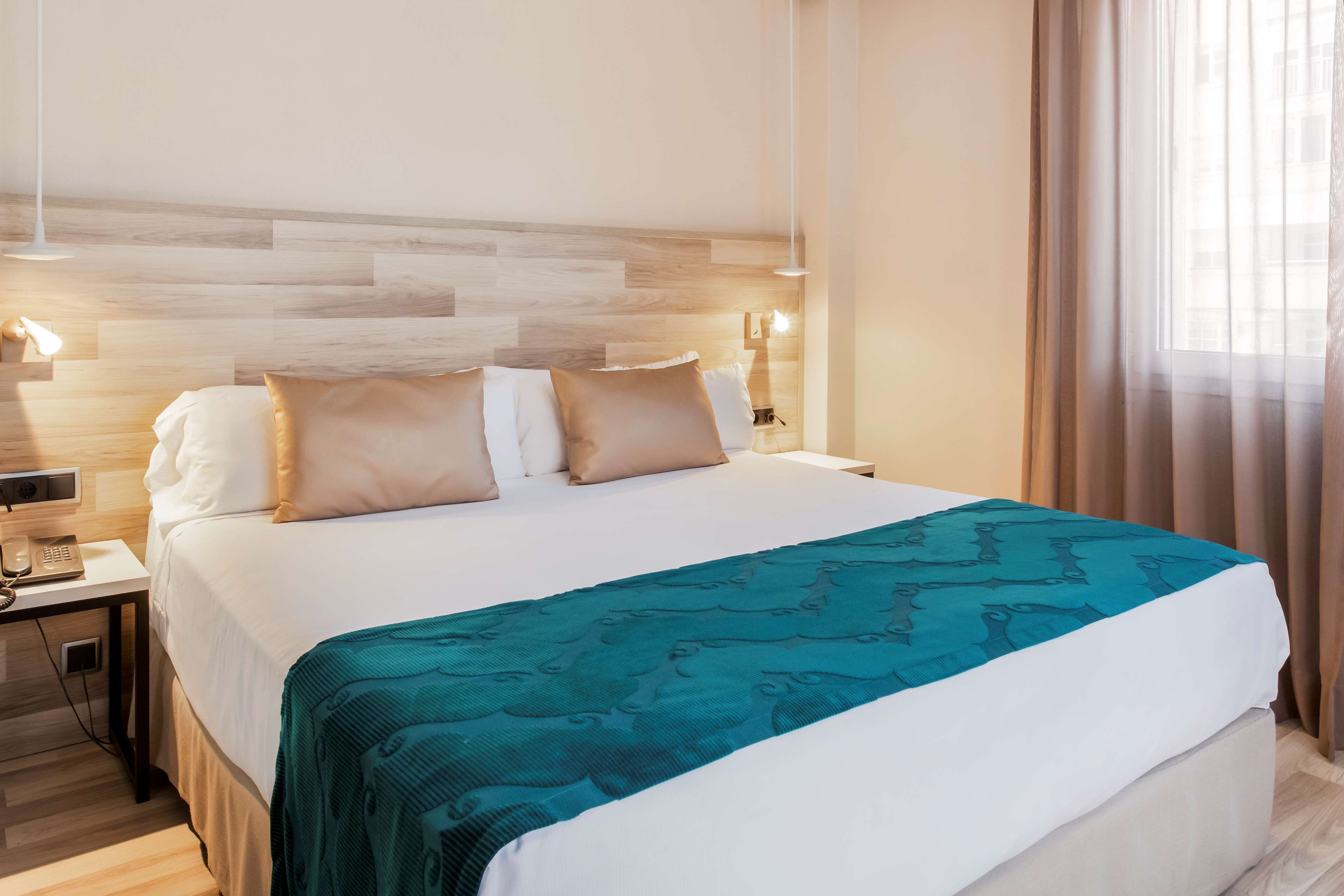 Quarto de hotel com uma cama grande, cabeceira de madeira, mesa de cabeceira com abajur e janela com cortinas.