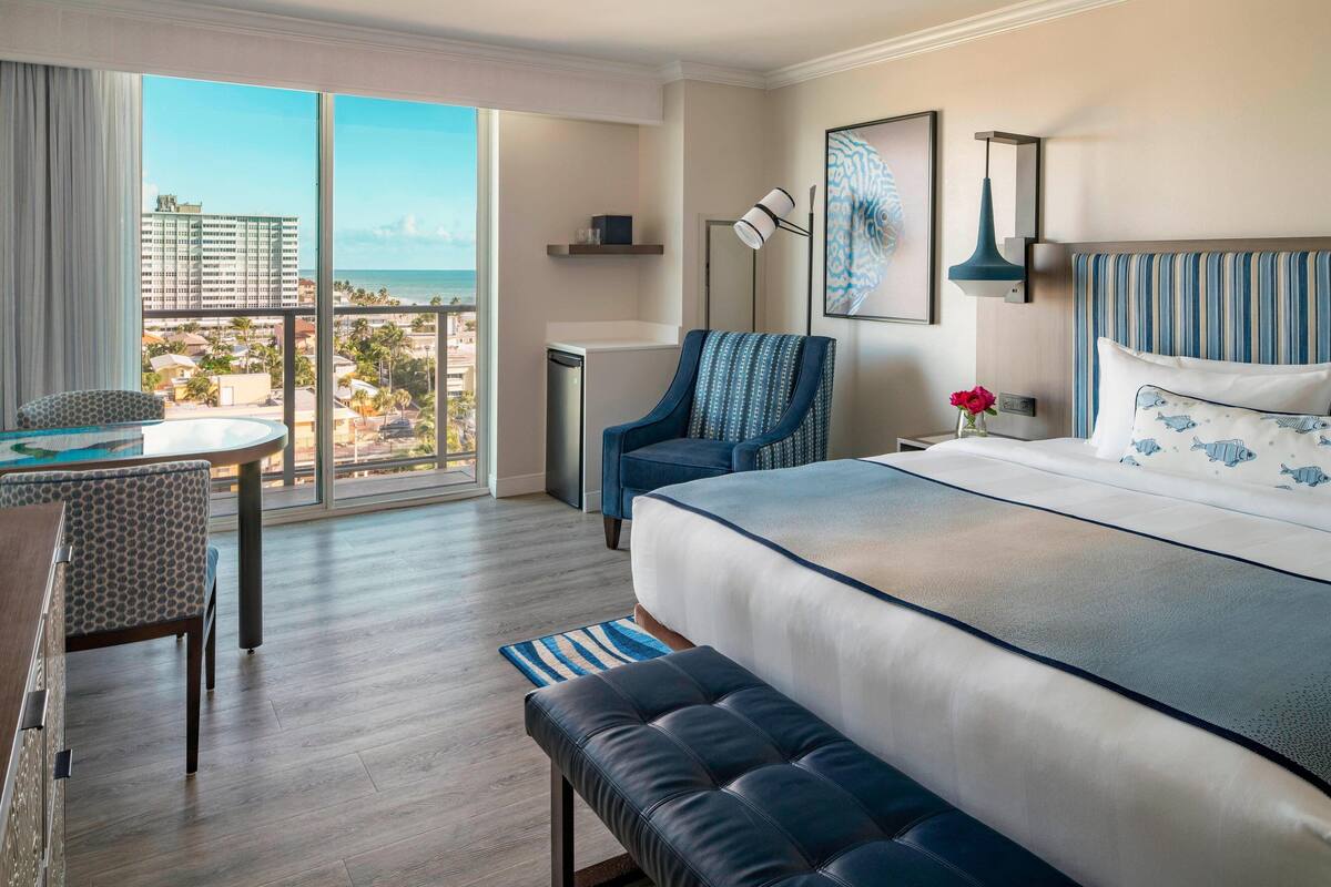 Quarto de hotel com uma cama grande, uma mesa, uma cadeira e vista para a cidade e oceano.