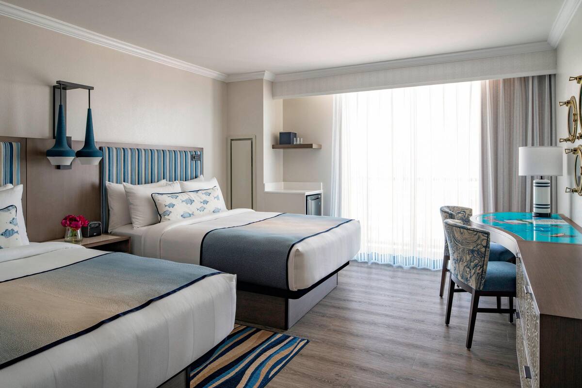 Quarto de hotel com duas camas, uma escrivaninha com cadeira, uma janela grande com cortinas e um minibar.