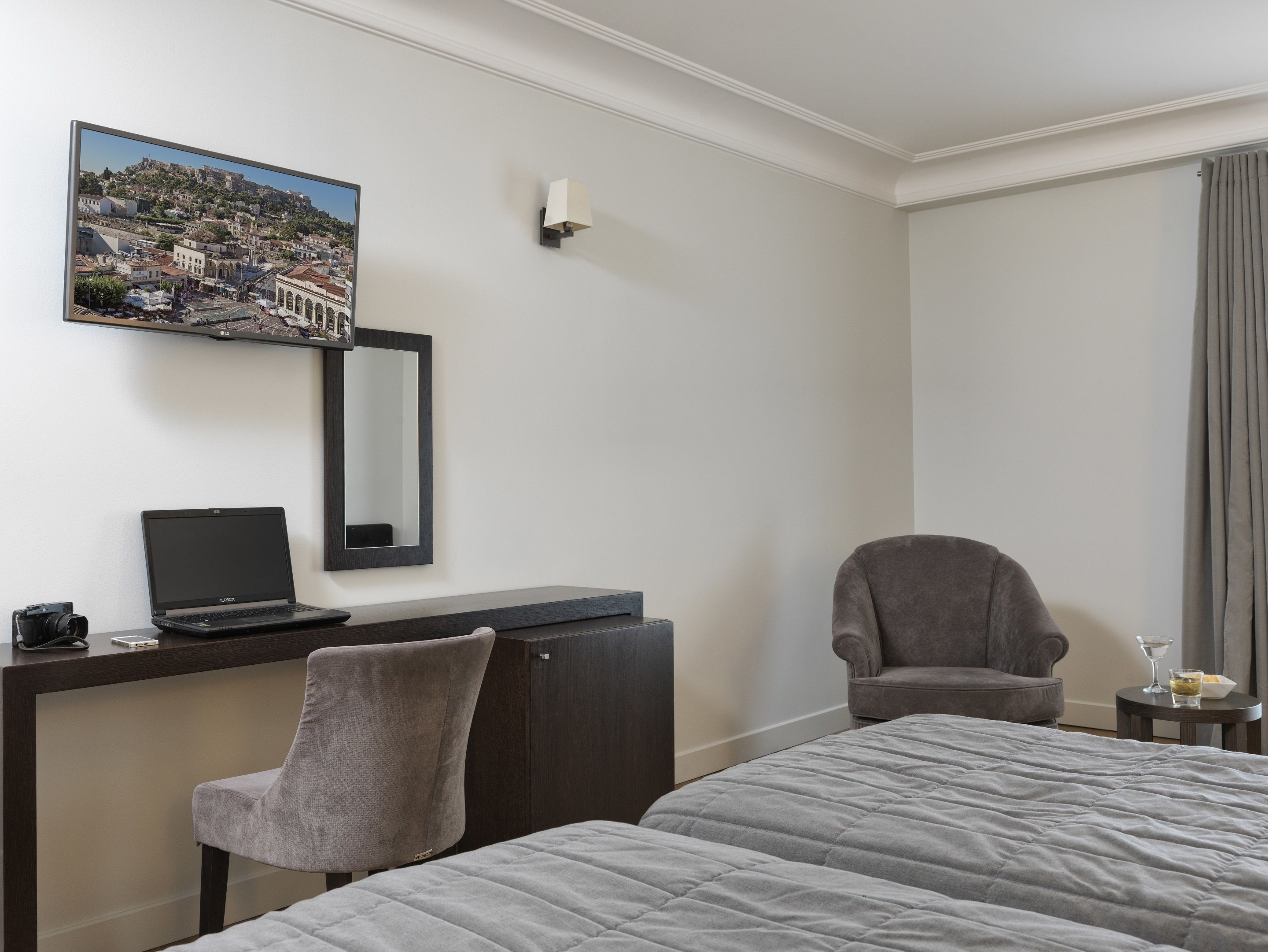 Quarto de hotel com uma cama, uma escrivaninha com um laptop, uma cadeira, um espelho e uma janela com cortinas.