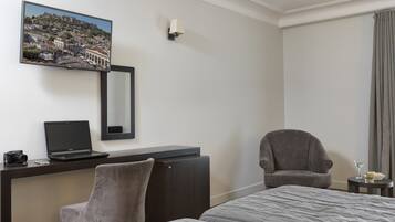 Quarto de hotel com uma cama, uma escrivaninha com um laptop, uma cadeira, um espelho e uma janela com cortinas.