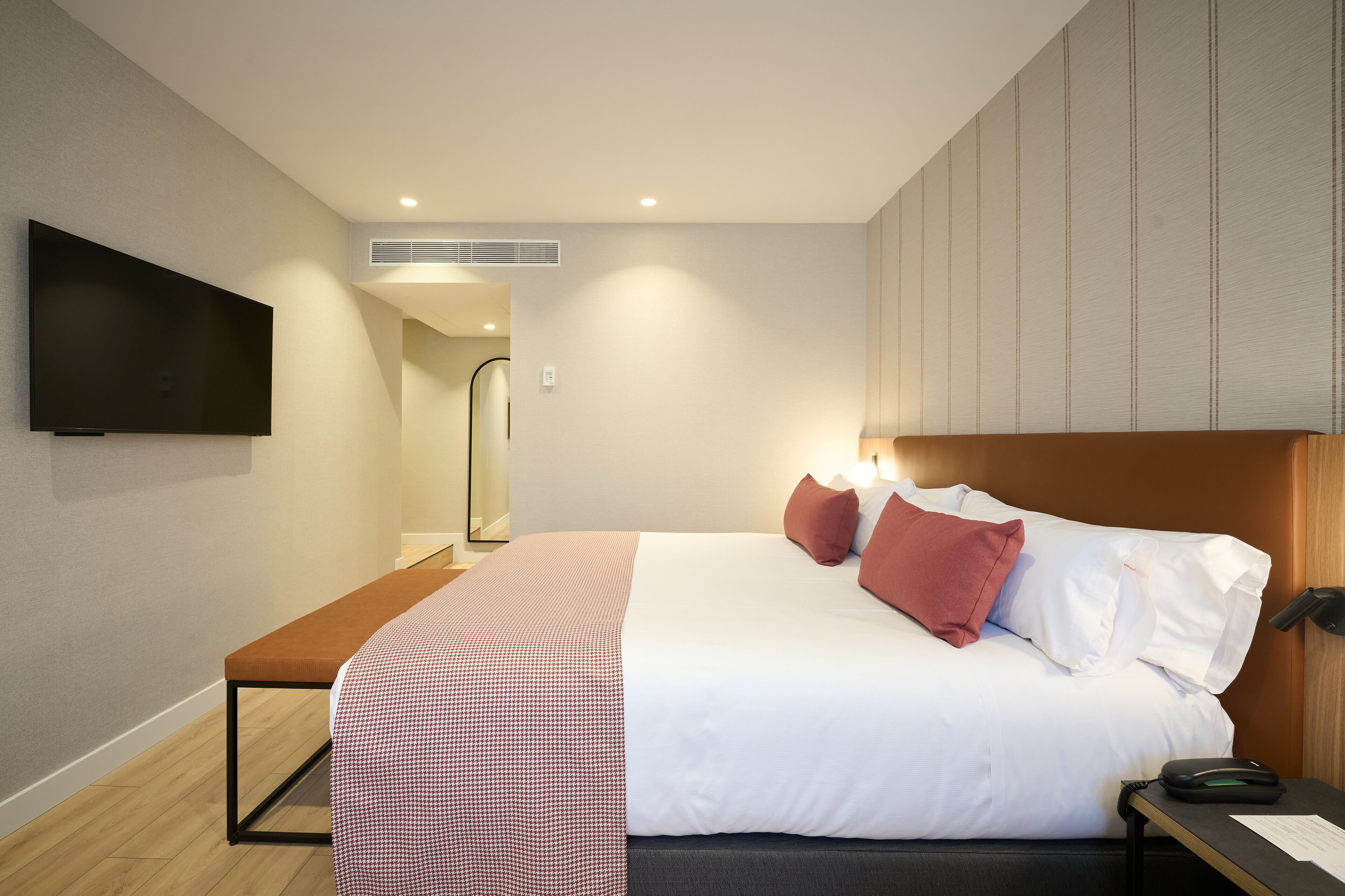 Quarto de hotel moderno com uma cama grande, televisão de tela plana na parede, mesa de cabeceira e um banco.