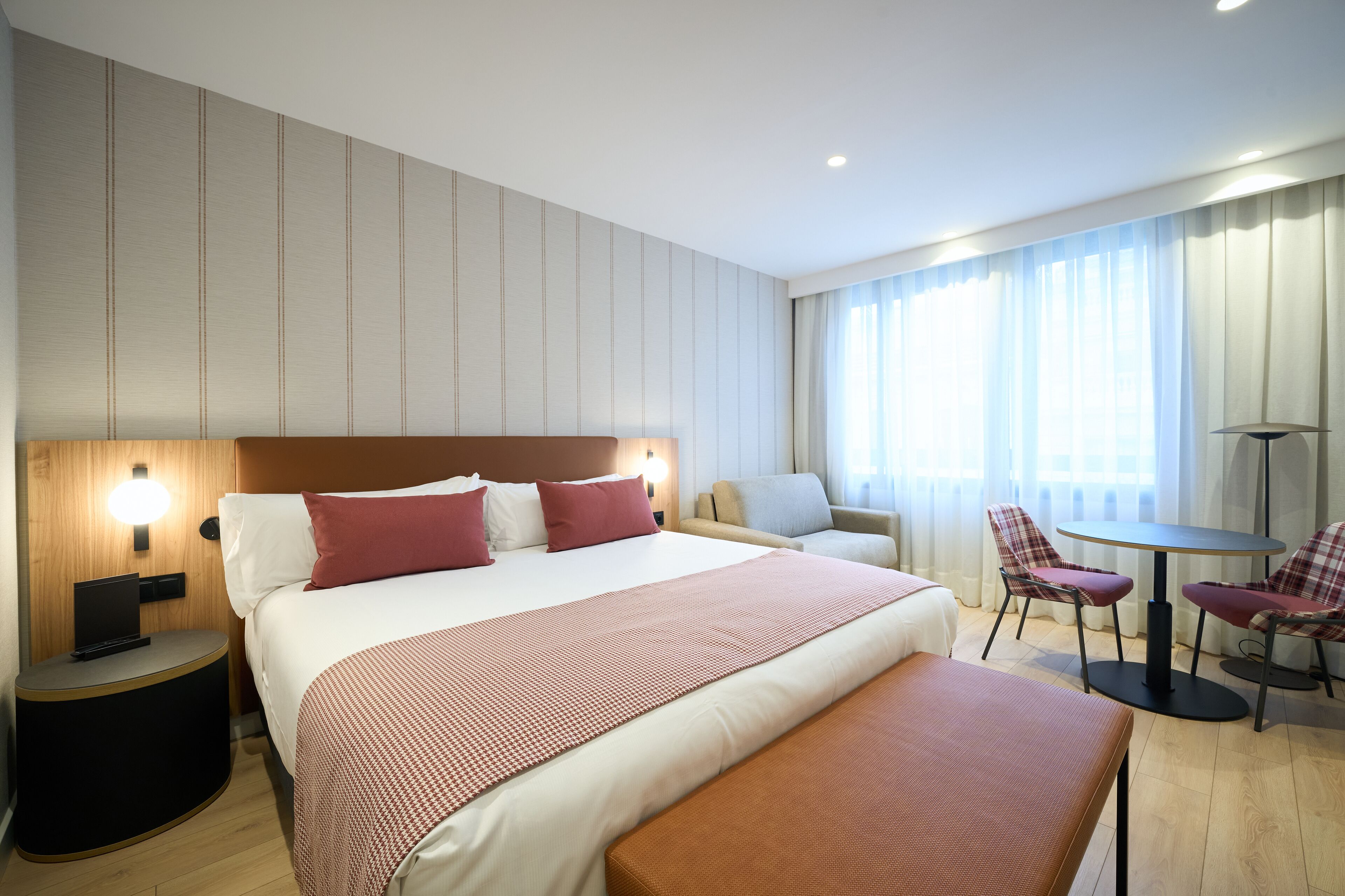 Quarto de hotel com uma cama grande, uma mesa de cabeceira com abajur, uma mesinha redonda com duas cadeiras e um sofá.