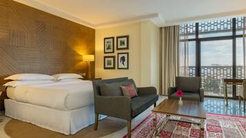 Quarto de hotel com cama, sofá, mesa de centro e vista da cidade por grandes janelas.