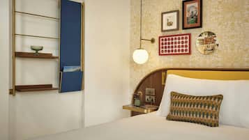 Quarto de hotel com cama, mesa de cabeceira, prateleira na parede com uma tigela, porta azul e arte decorativa na parede.