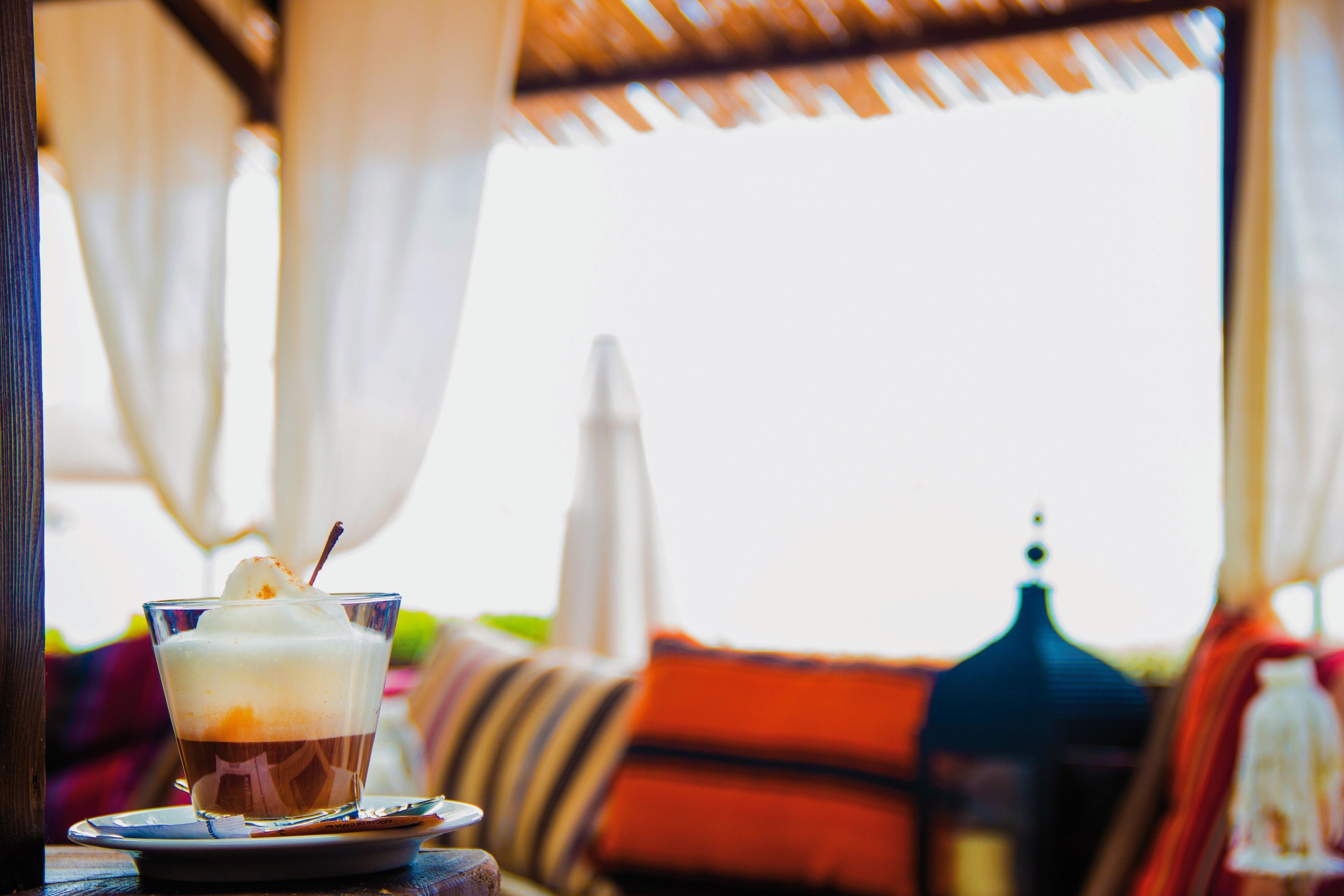 Um copo de café gelado com uma colher num pires, um prato e uma almofada listrada, num ambiente acolhedor ao ar livre.