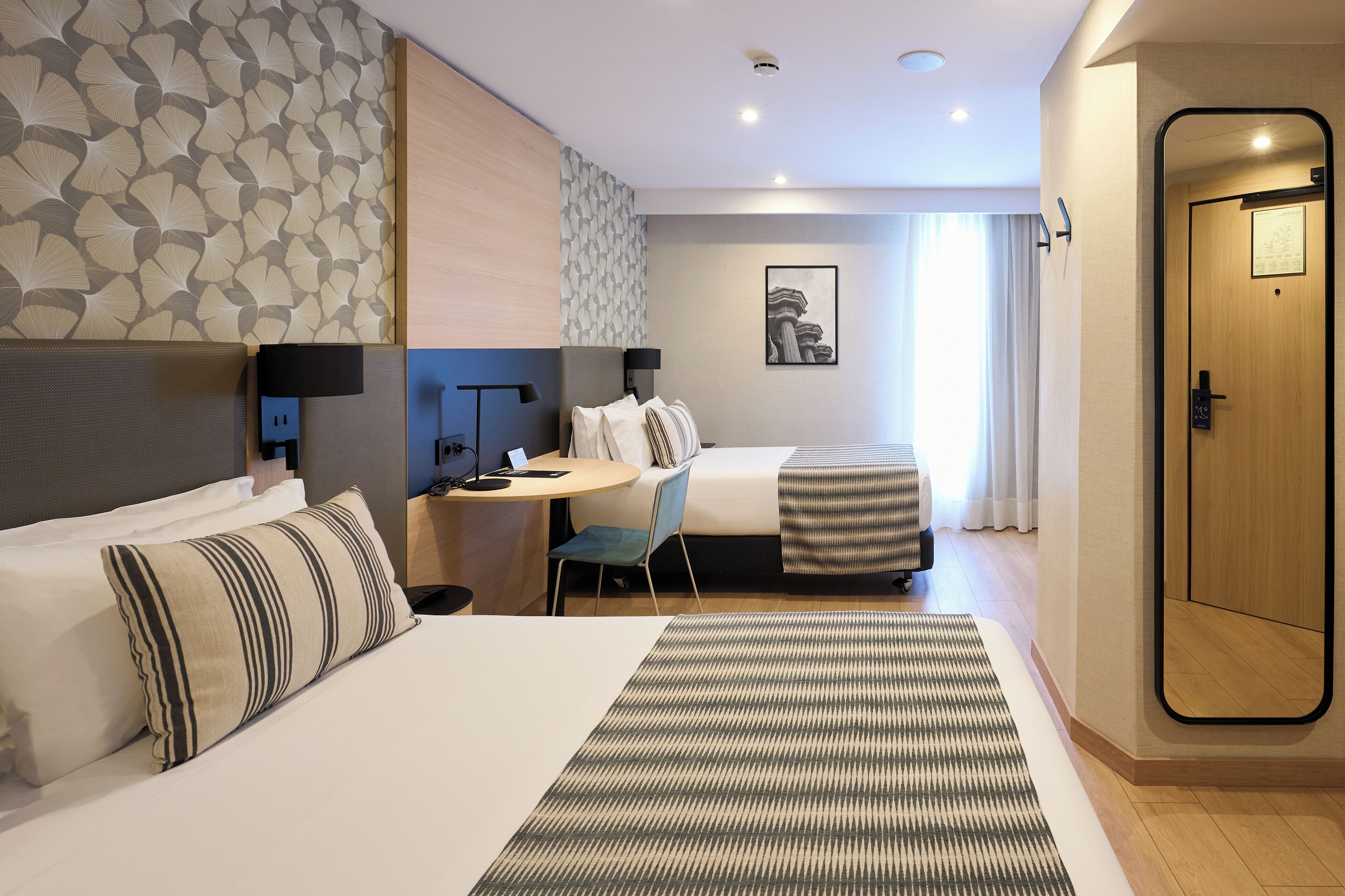 Quarto de hotel com cama, escrivaninha, cadeira, mesinha e porta.