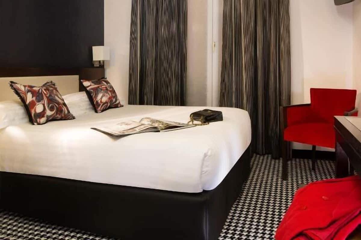 Quarto de hotel com uma cama, uma cadeira vermelha, uma mesa e um vaso com flores.