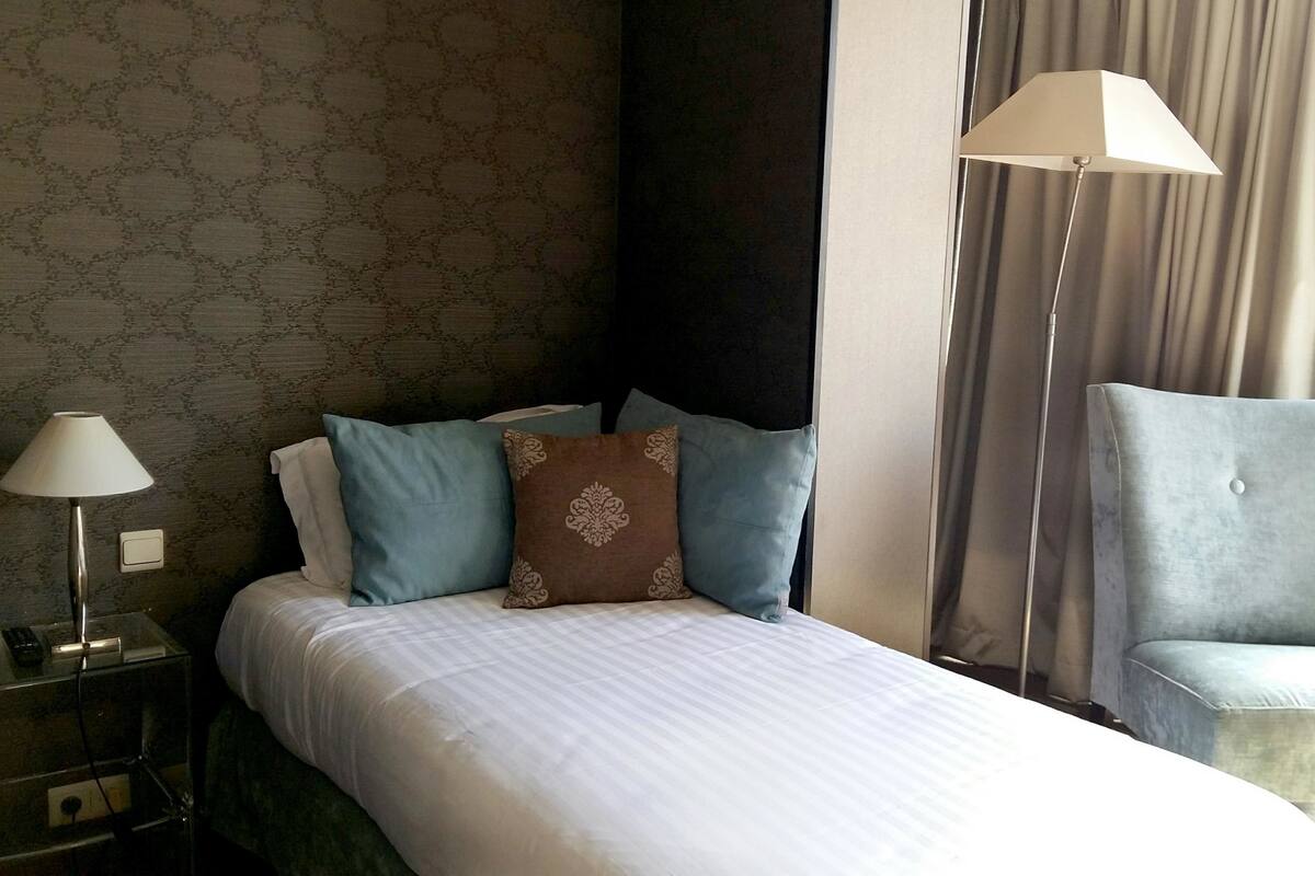 Quarto de hotel com uma cama bem arrumada, mesa de cabeceira com abajur e uma cadeira.