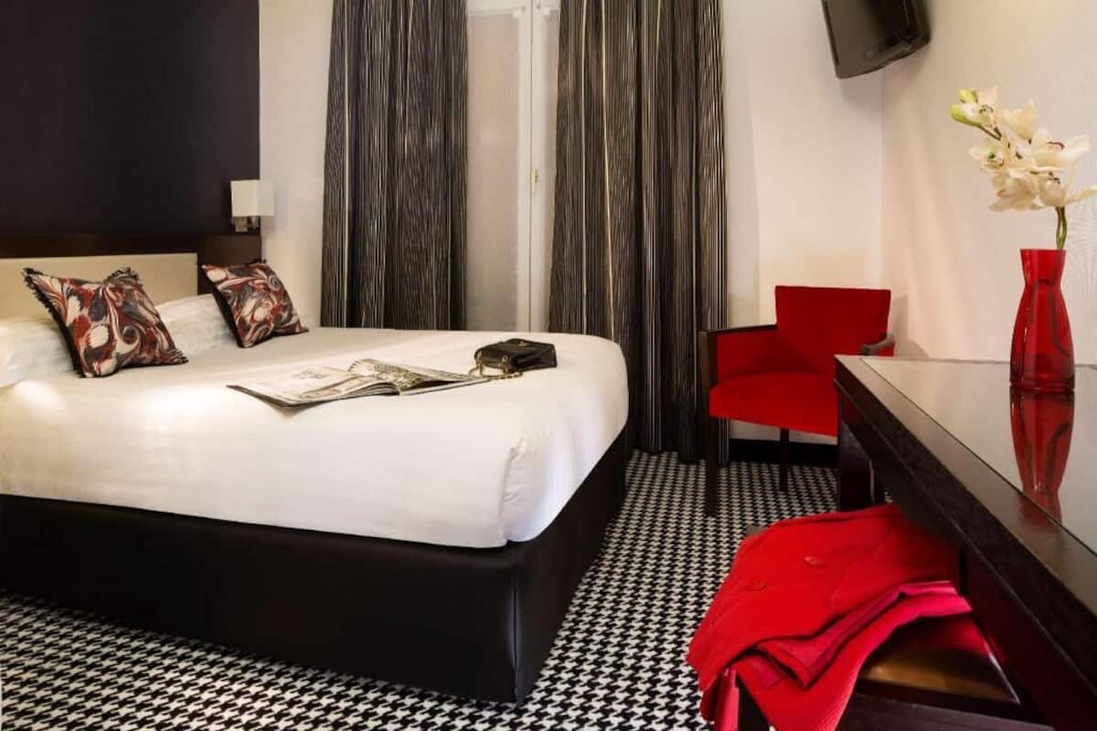 Quarto de hotel com uma cama, uma cadeira vermelha, um vaso vermelho com flores e uma televisão na parede.