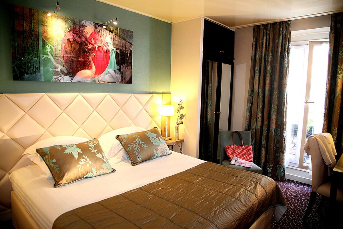Quarto de hotel com uma cama grande, cabeceira, um quadro na parede, mesa de cabeceira com abajur e vista para o exterior através de uma porta.