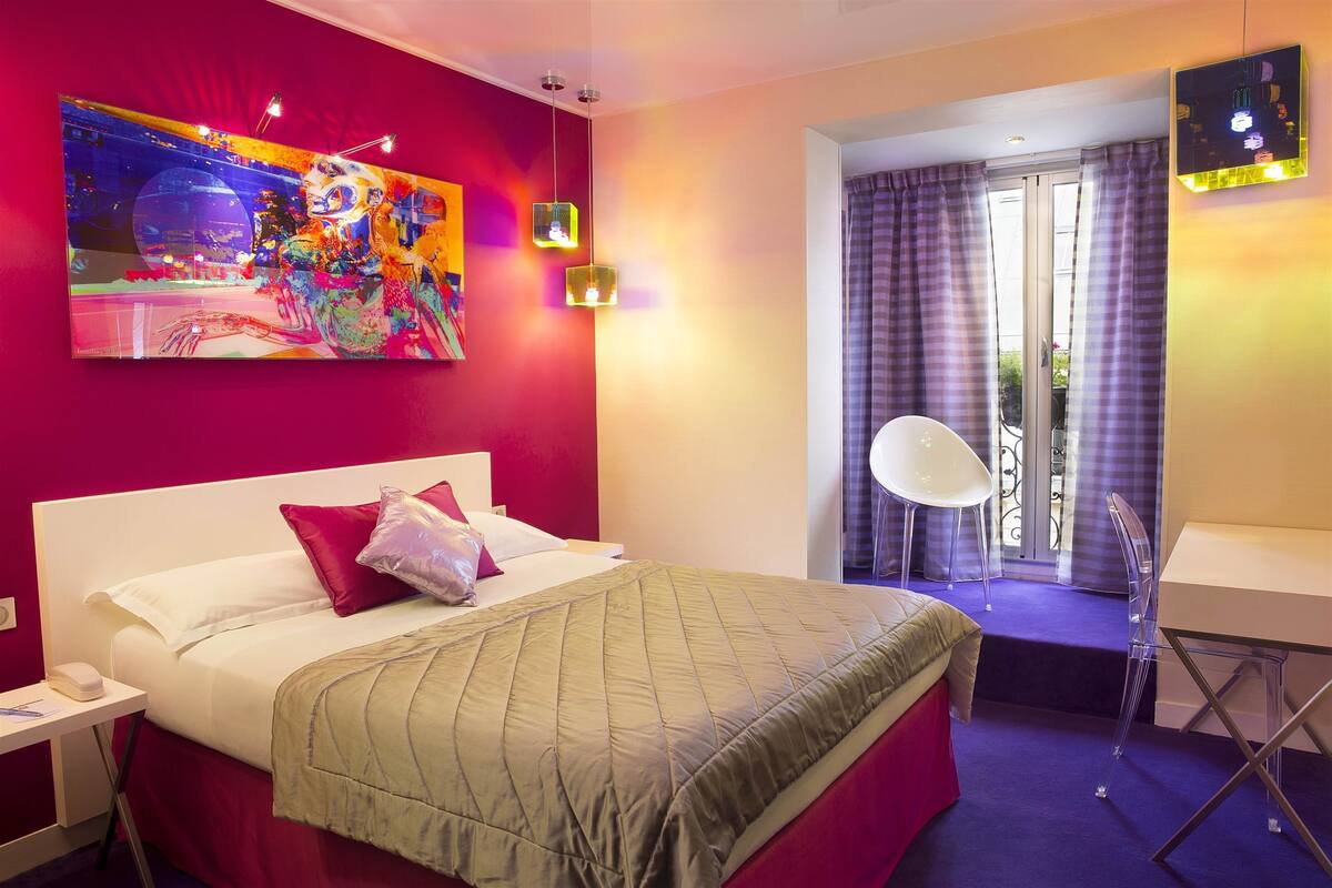 Quarto de hotel com uma cama, uma secretária, uma cadeira e uma janela grande com cortinas.