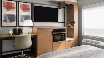 Quarto de hotel com escrivaninha de madeira, uma cadeira moderna, cama com roupa de cama listrada, televisão de tela plana, micro-ondas, cafeteira e janela com persianas.