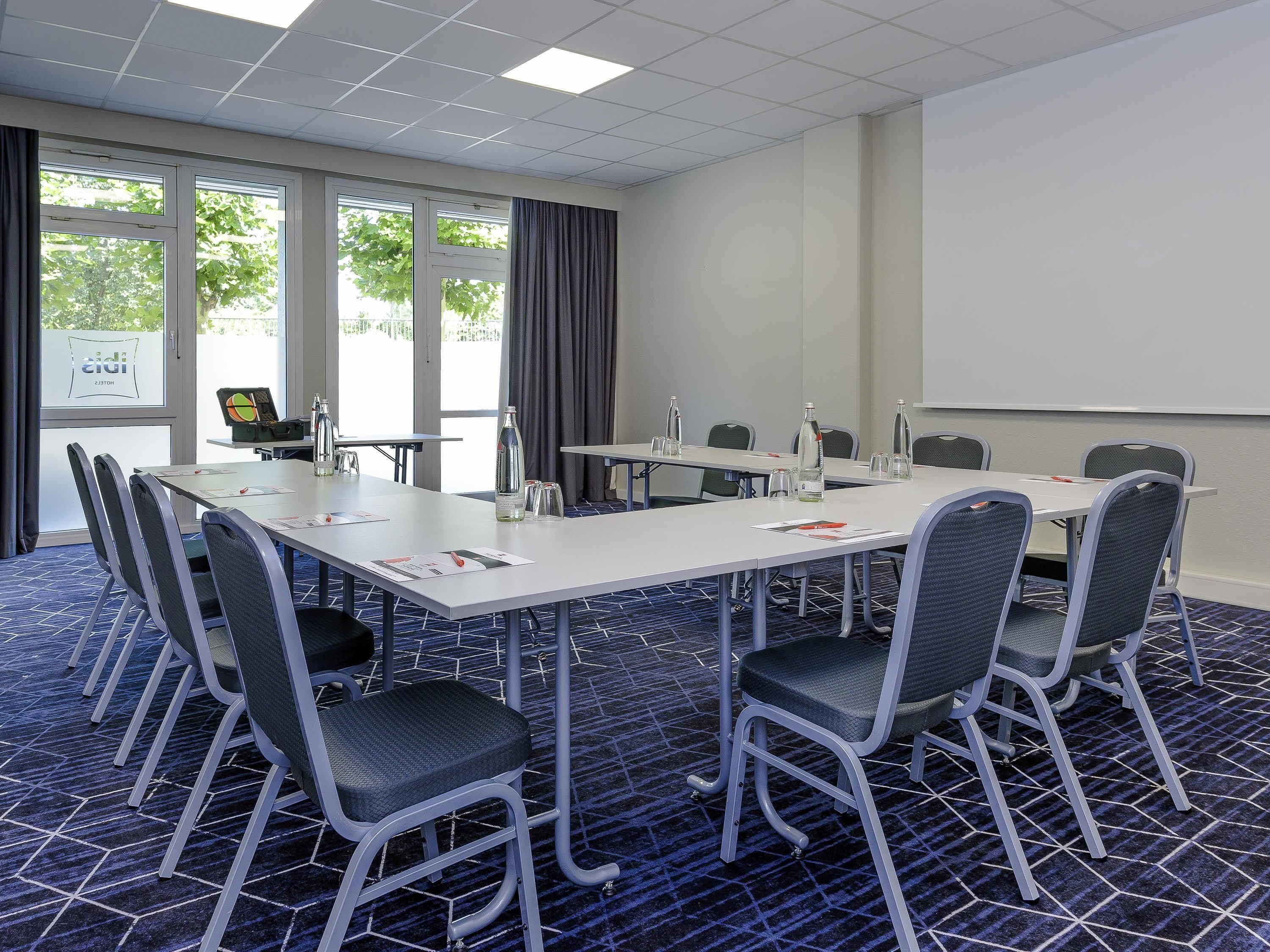 Uma sala de conferência com uma mesa comprida, cadeiras, uma tela de projetor e uma janela com cortinas.