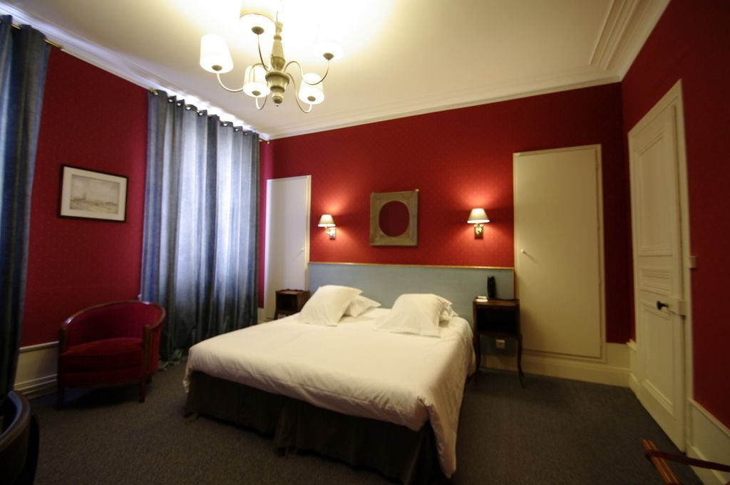 Quarto de hotel com uma cama grande, parede vermelha, lustre e duas luminárias de cabeceira.