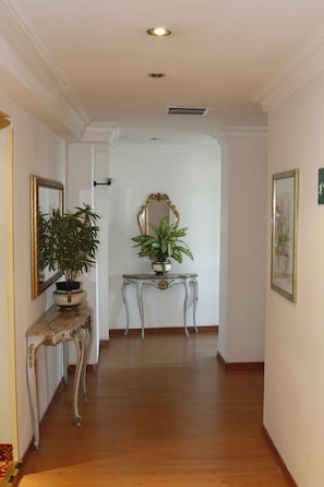 Um corredor com piso de madeira, parede branca e porta branca no final. Há duas plantas em vasos sobre uma mesa consola branca.