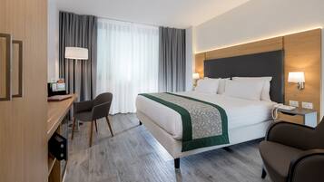 Quarto de hotel com cama, mesa de cabeceira, abajur, escrivaninha, cadeira e janela com cortinas.