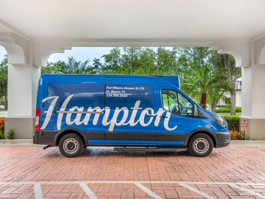 Uma carrinha de entregas azul da Hampton estacionada sob uma cobertura, com pavimento de tijolo, rodeada por vegetação e um aviso de 'Proibido Estacionar'.