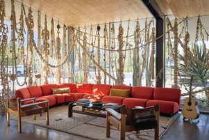 Uma sala de estar moderna com um sofá vermelho, mesa de centro de madeira e cadeiras, decorada com peças de macramé nas paredes e um violão.