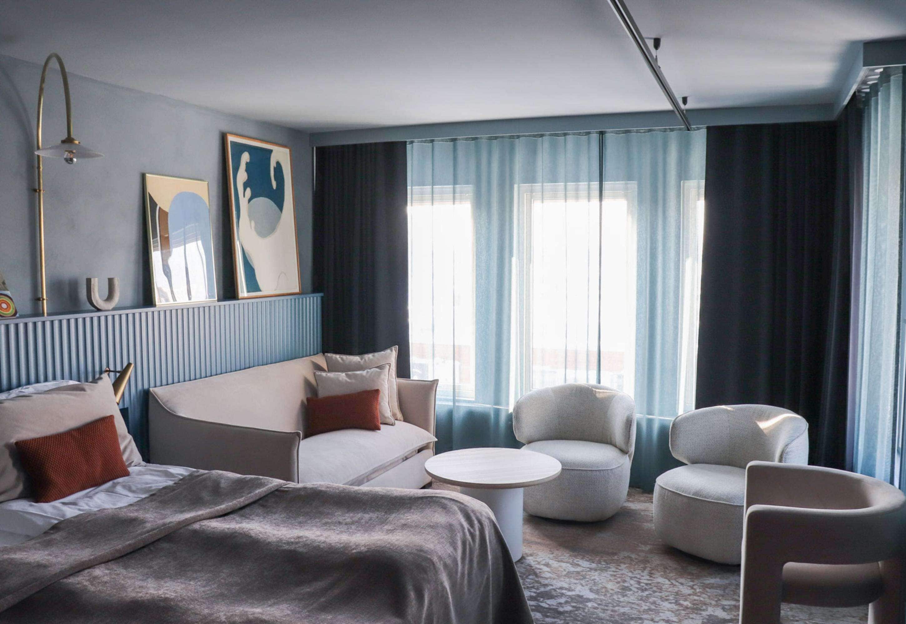 Quarto de hotel moderno com uma cama, duas poltronas, uma mesa de centro redonda e uma janela grande com cortinas.