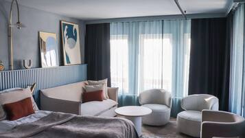 Quarto de hotel moderno com uma cama, duas poltronas, uma mesa de centro redonda e uma janela grande com cortinas.
