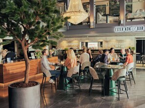 Uma área de bar moderna em ambiente interno, com mesas e cadeiras redondas, um balcão de recepção e um grande candelabro.