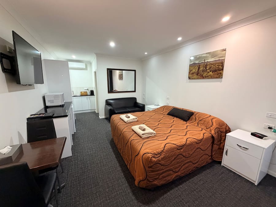 Quarto de hotel com cama, escrivaninha, cadeira, televisão, quadro e mesa pequena.