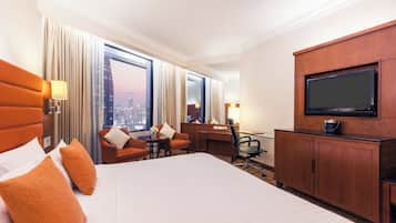 Quarto de hotel com uma cama grande, uma escrivaninha, uma televisão e vista para a paisagem urbana pelas janelas.