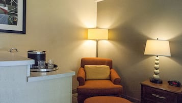 Quarto de hotel com uma pequena cozinha, sofá, mesa lateral com luminária e um quadro na parede.