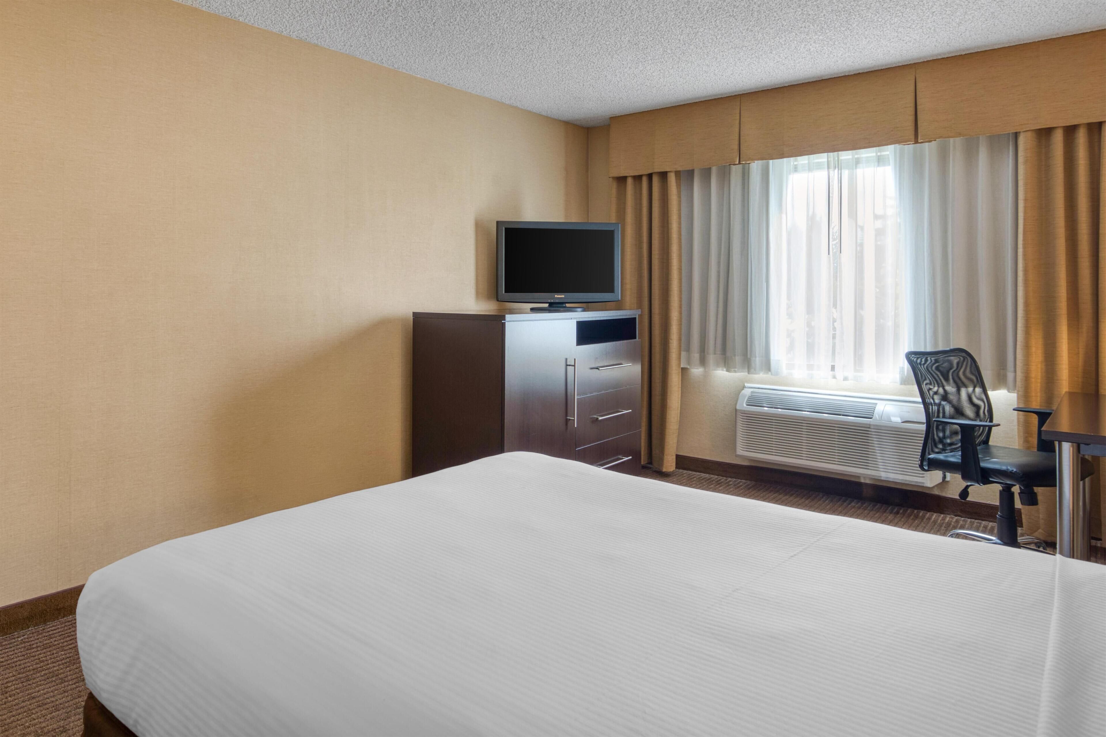 Quarto de hotel com cama, televisão em uma cômoda, escrivaninha com cadeira e unidade de ar condicionado.