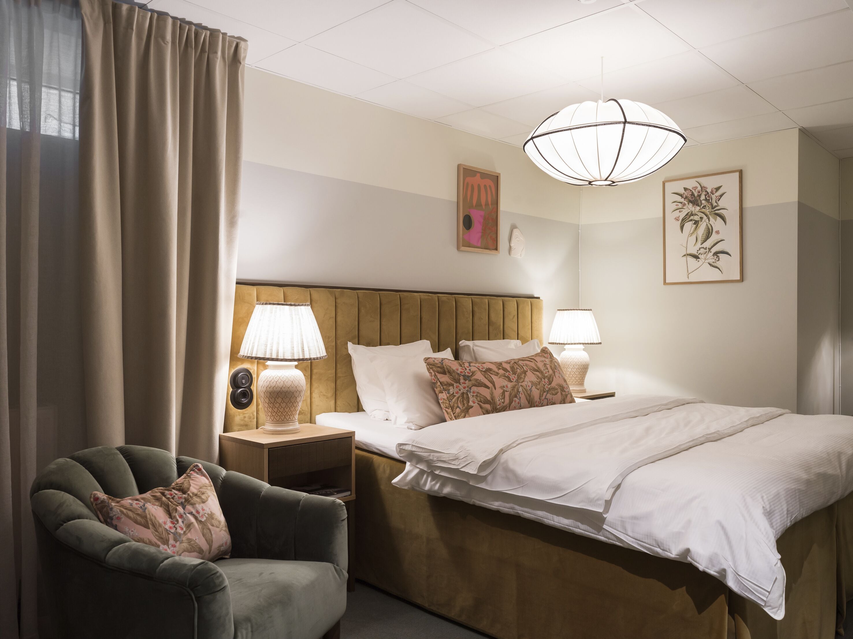 Quarto de hotel com cama, duas luminárias de cabeceira, mesa de cabeceira, cadeira e uma grande janela com cortinas.